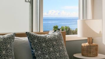 Quarto com cama, almofadas estampadas, mesa de cabeceira com abajur e vista para o mar pela janela.