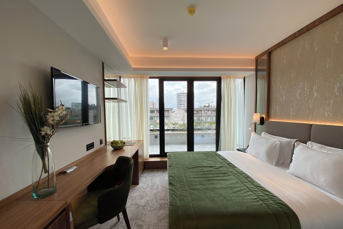 Quarto de hotel com cama, escrivaninha, cadeira, televisão e vista para edifícios pela janela.