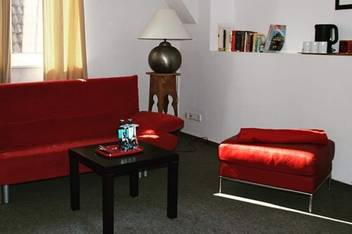 Uma sala de estar com um sofá vermelho, uma poltrona vermelha, uma mesinha, um abajur, uma estante de livros e uma máquina de café.