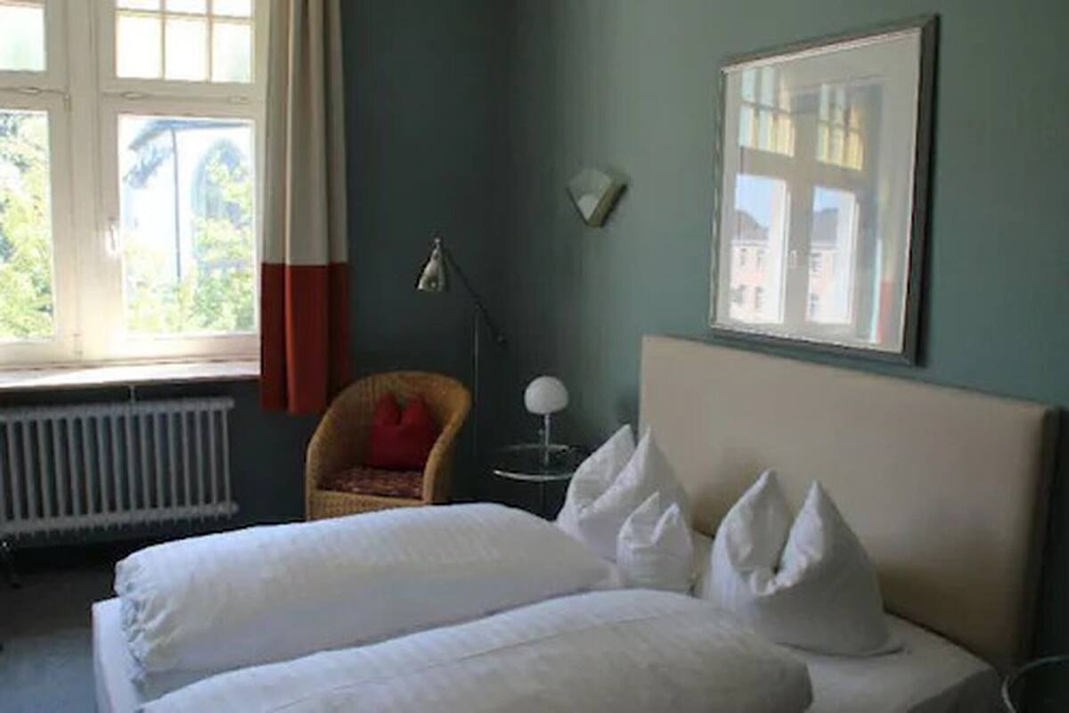 Quarto de hotel com uma cama, uma cadeira, um abajur e uma janela com cortinas.