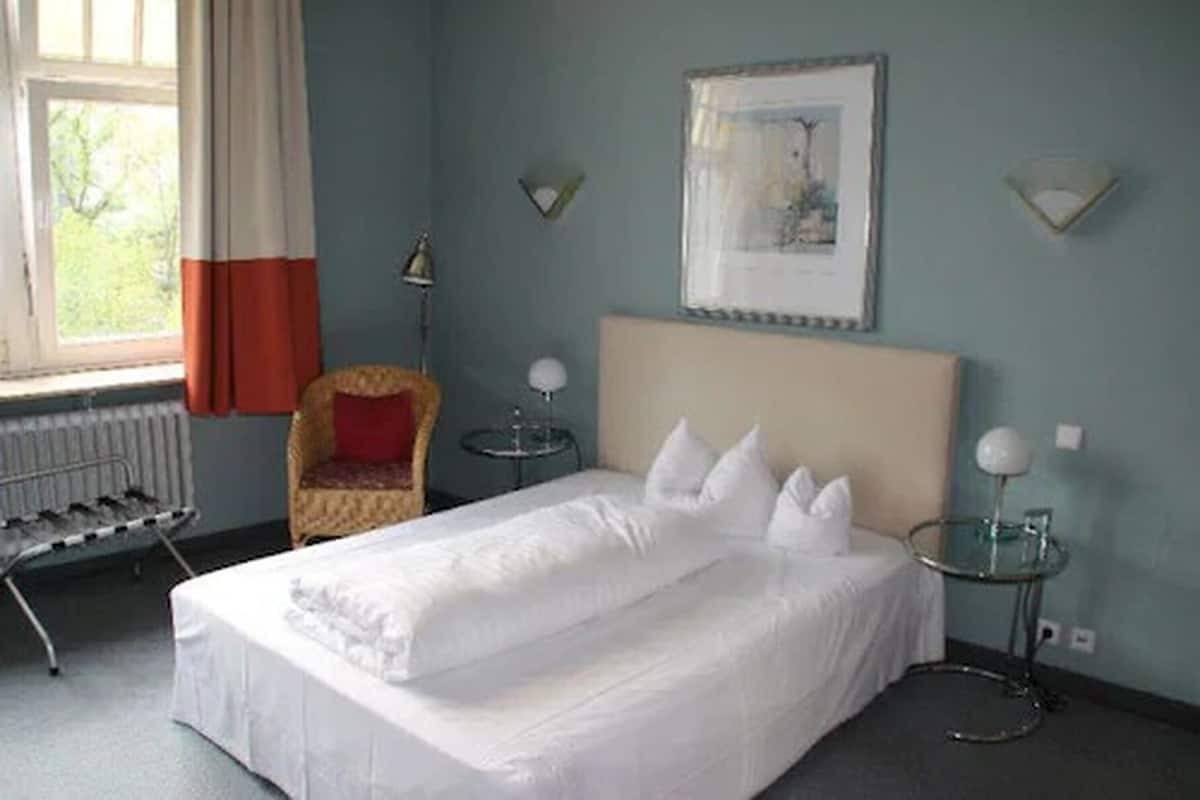 Uma cama bem arrumada com lençóis brancos em um quarto com parede azul, um quadro e uma pequena mesa redonda com abajur.