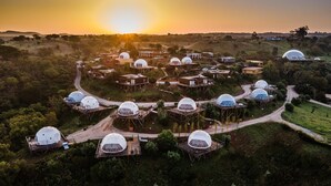 Conjunto de acomodações em formato de cúpula, erguidas sobre pilares, emolduradas por uma paisagem natural com o pôr do sol ao fundo.