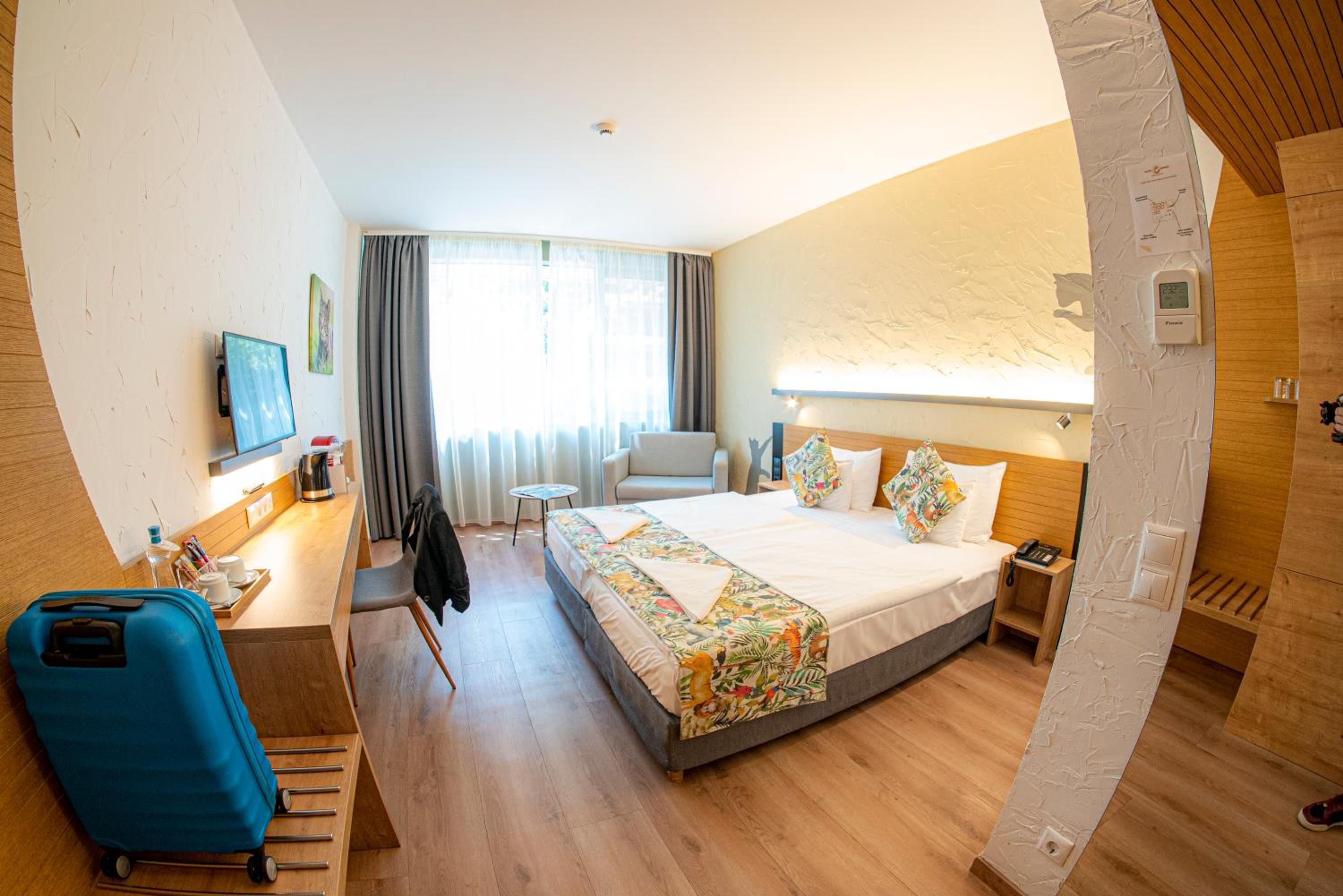 Quarto de hotel com cama, escrivaninha, cadeira, mala e janela com cortinas.
