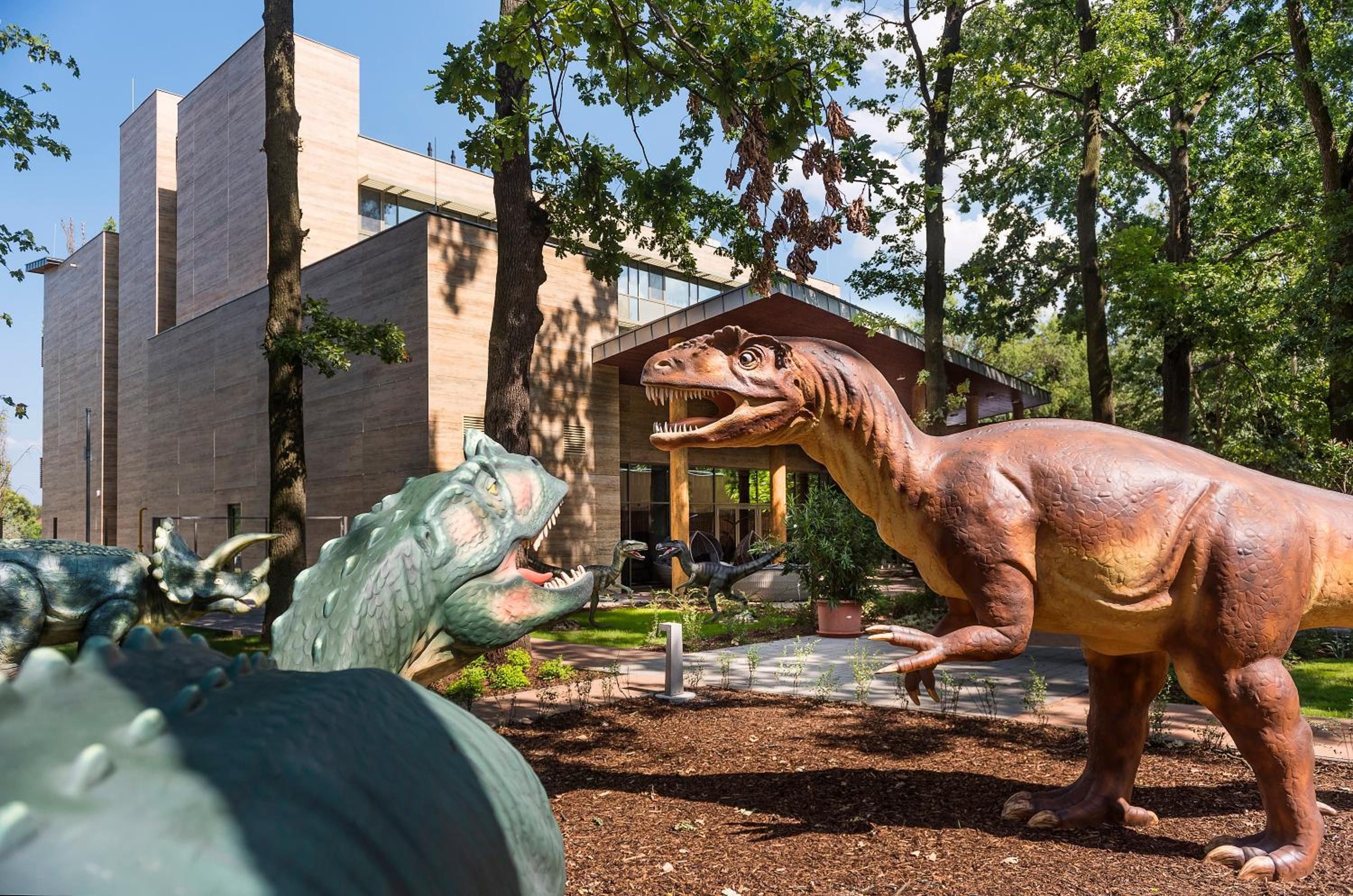Exposição de dinossauros com um grande Tiranossauro Rex e um Estegossauro em frente a um edifício moderno com entrada coberta.