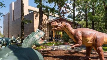 Exposição de dinossauros com um grande Tiranossauro Rex e um Estegossauro em frente a um edifício moderno com entrada coberta.