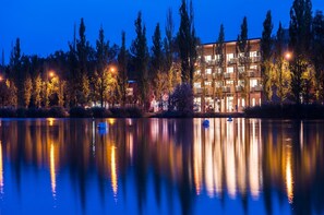 Um hotel com vários andares, varandas, um lago em primeiro plano e árvores ao fundo.
