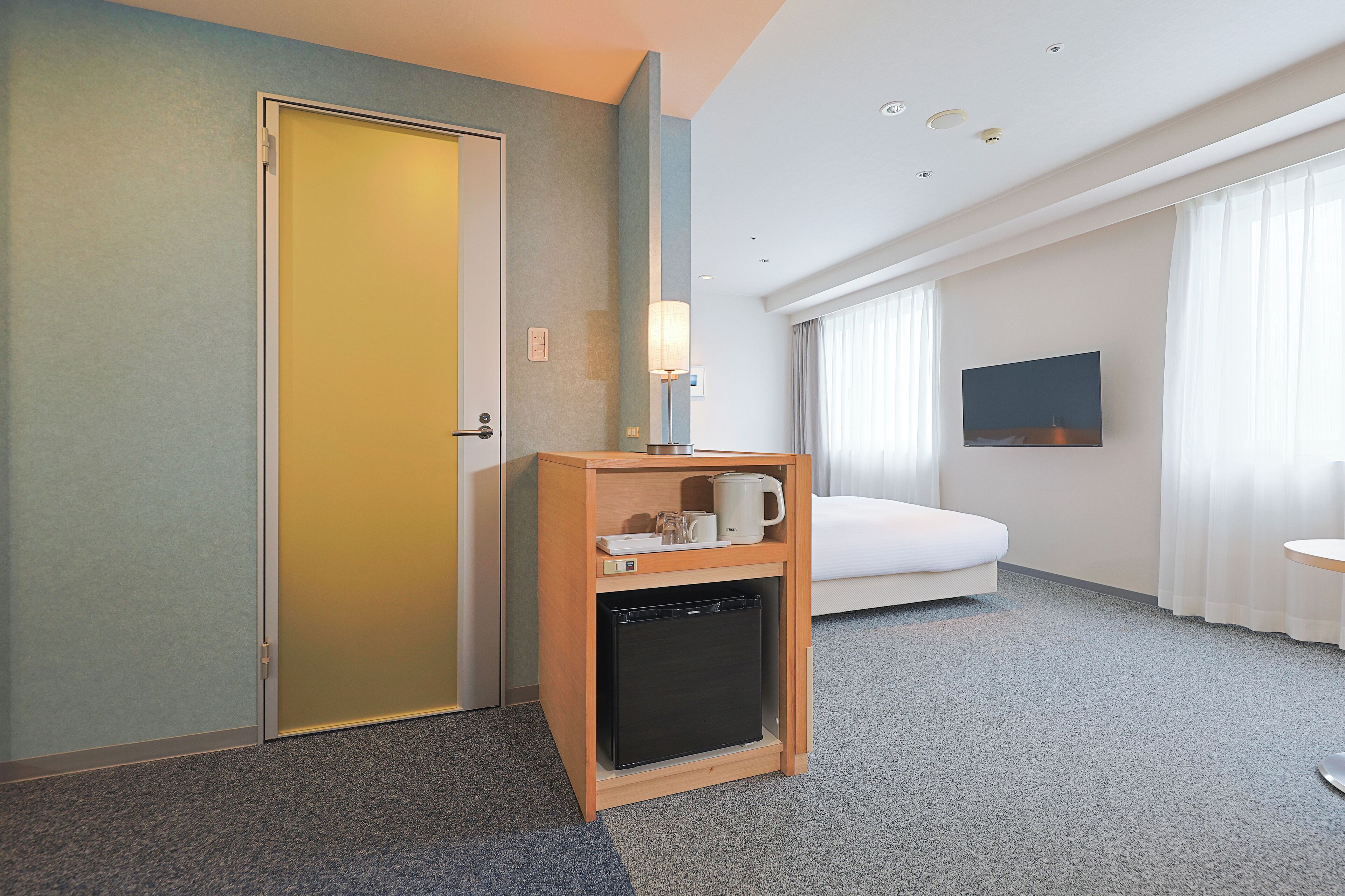 Quarto de hotel com porta, pequeno armário de madeira com cafeteira, sofá branco e TV de tela plana na parede.
