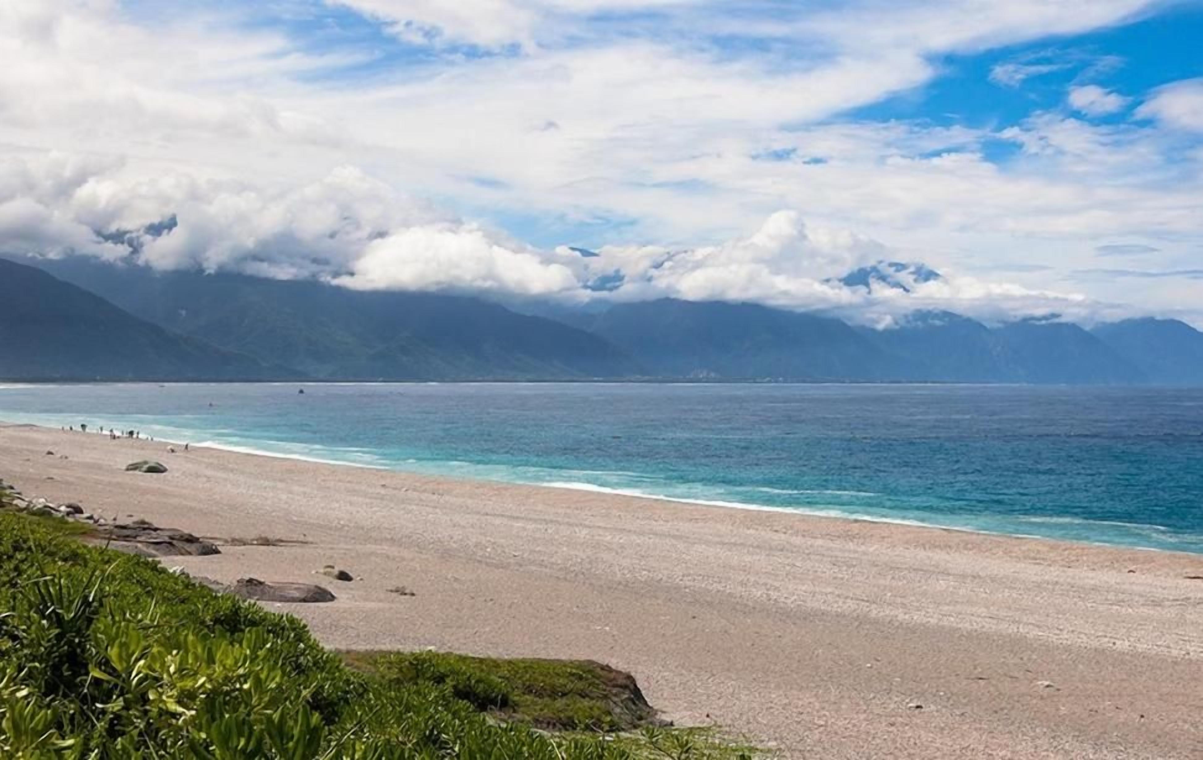 Uma praia com céu claro, montanhas ao fundo e mar calmo.