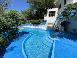 Uma piscina com água azul cristalina, rodeada por árvores e um edifício branco com escadaria de madeira.