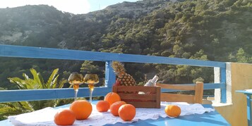 Mesa com laranjas e dois copos de vinho sobre toalha azul, com vista para uma paisagem montanhosa.