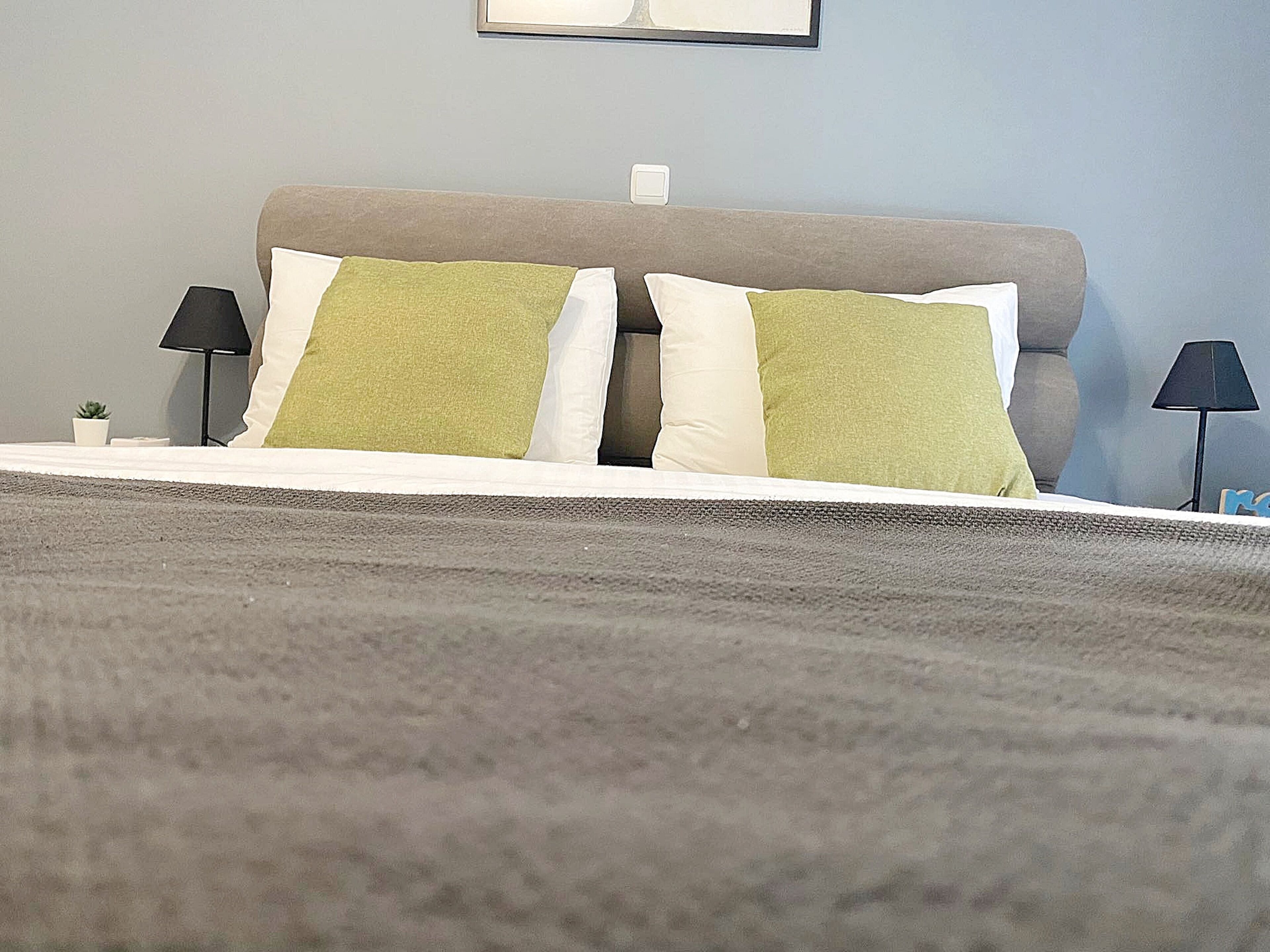 Uma cama bem arrumada com cabeceira cinza, duas almofadas e um cobertor cinza. Há duas mesas de cabeceira, cada uma com uma luminária e uma pequena planta em vaso.