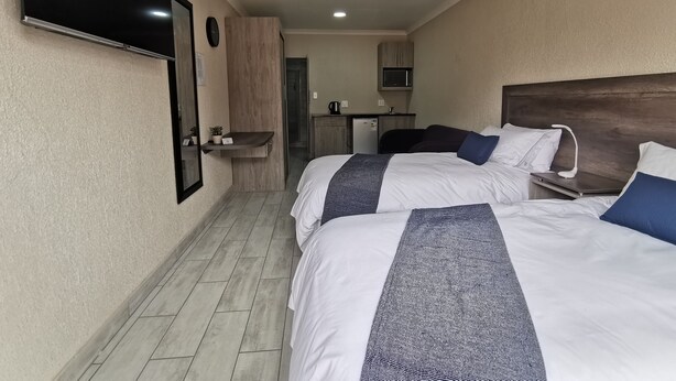 Quarto de hotel com duas camas, televisão, um pequeno escritório e uma cozinha compacta ao fundo.