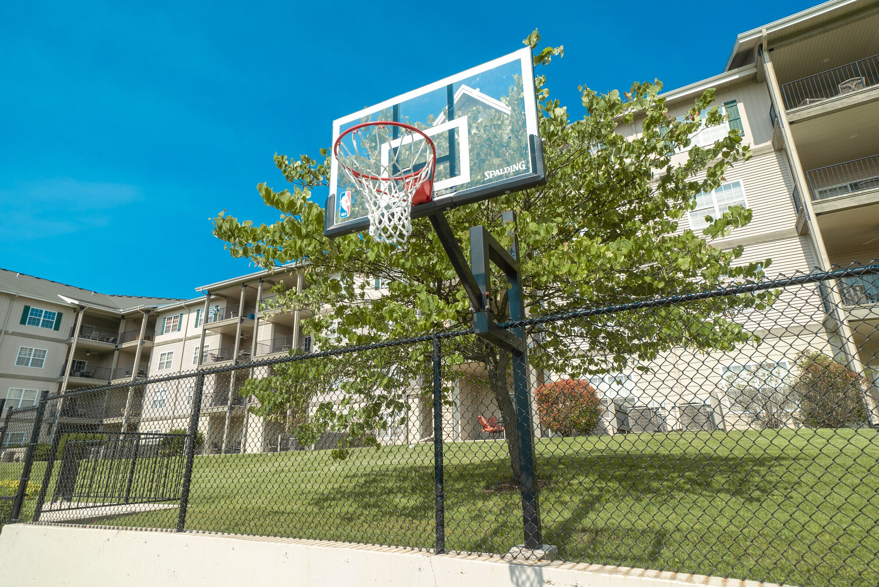 Uma quadra de basquete com um aro Spalding, cercada por uma cerca de arame, em frente a um prédio com varandas.
