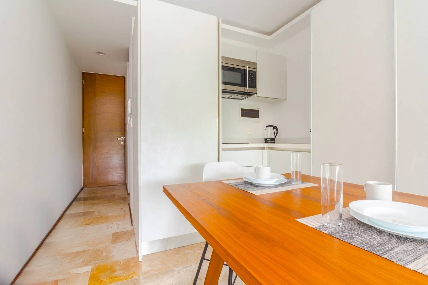 Uma cozinha compacta com armários brancos e um micro-ondas, uma área de refeições com mesa e cadeiras de madeira, e um corredor que dá para outro quarto.