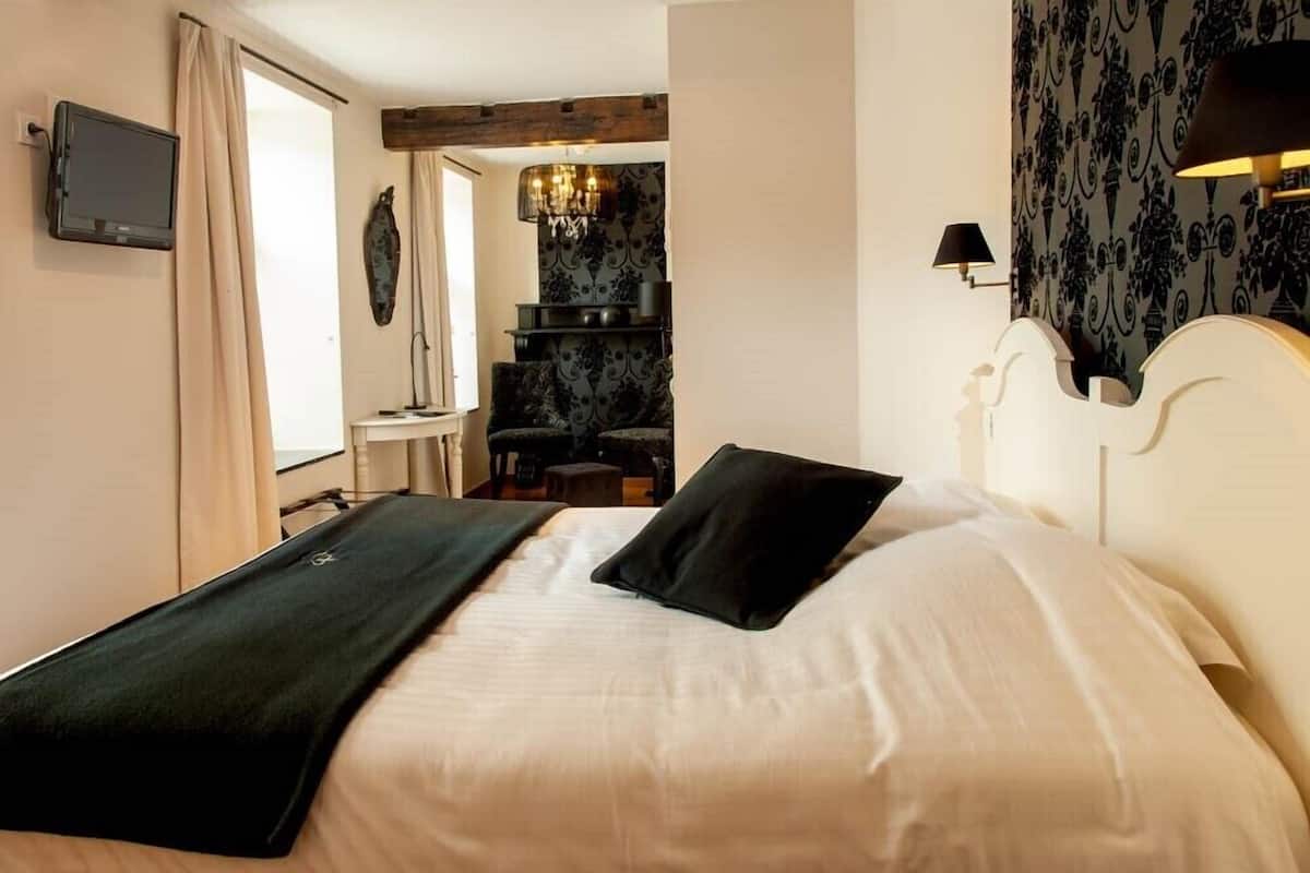 Cama bem arrumada com travesseiro preto e edredom branco, televisão na parede, lustre e papel de parede estampado.