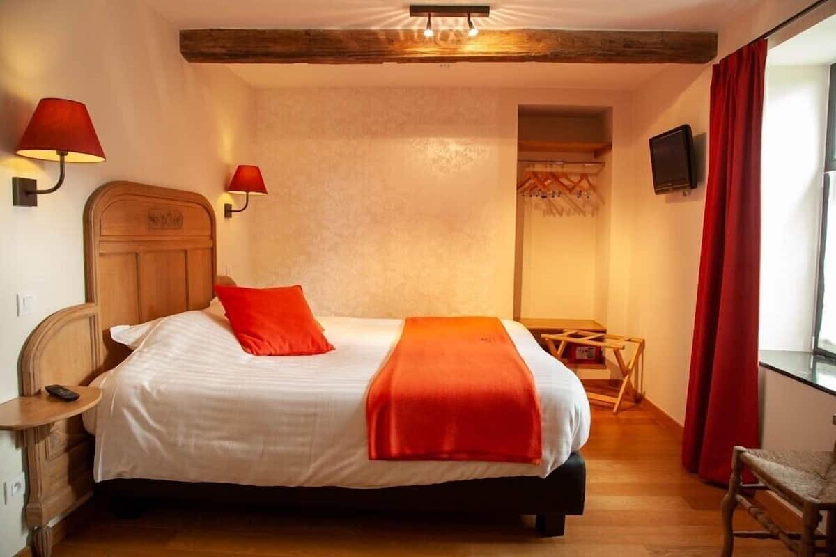 Quarto com cabeceira de madeira, cama com roupa de cama branca e detalhes vermelhos, duas luminárias de parede, uma televisão na parede e uma janela com cortinas vermelhas.