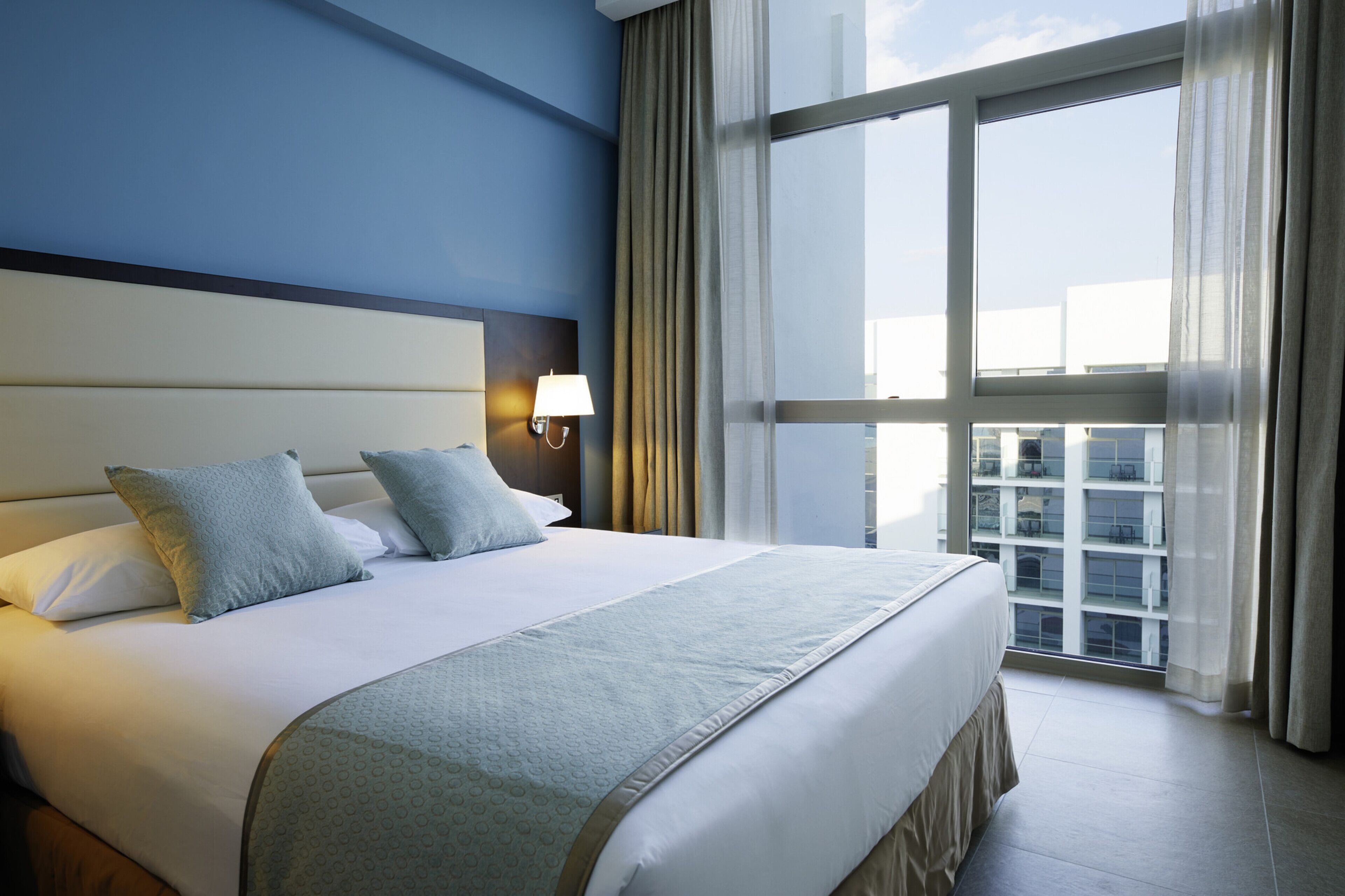 Quarto de hotel com uma cama grande, uma mesa de cabeceira com abajur e vista para edifícios pela janela.