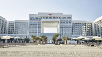 Um hotel moderno com um grande letreiro “RIU”, uma área de praia arenosa com espreguiçadeiras e guarda-sóis, e uma fonte central.