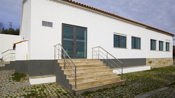 Um edifício moderno com fachada branca, um conjunto de escadas de madeira que levam a uma porta de vidro e uma pequena área ajardinada com um caminho de pedra.