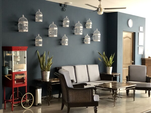 Uma sala de estar com parede azul, um sofá, duas poltronas, uma mesa de centro, uma máquina de vendas vermelha e vários vasos de plantas.