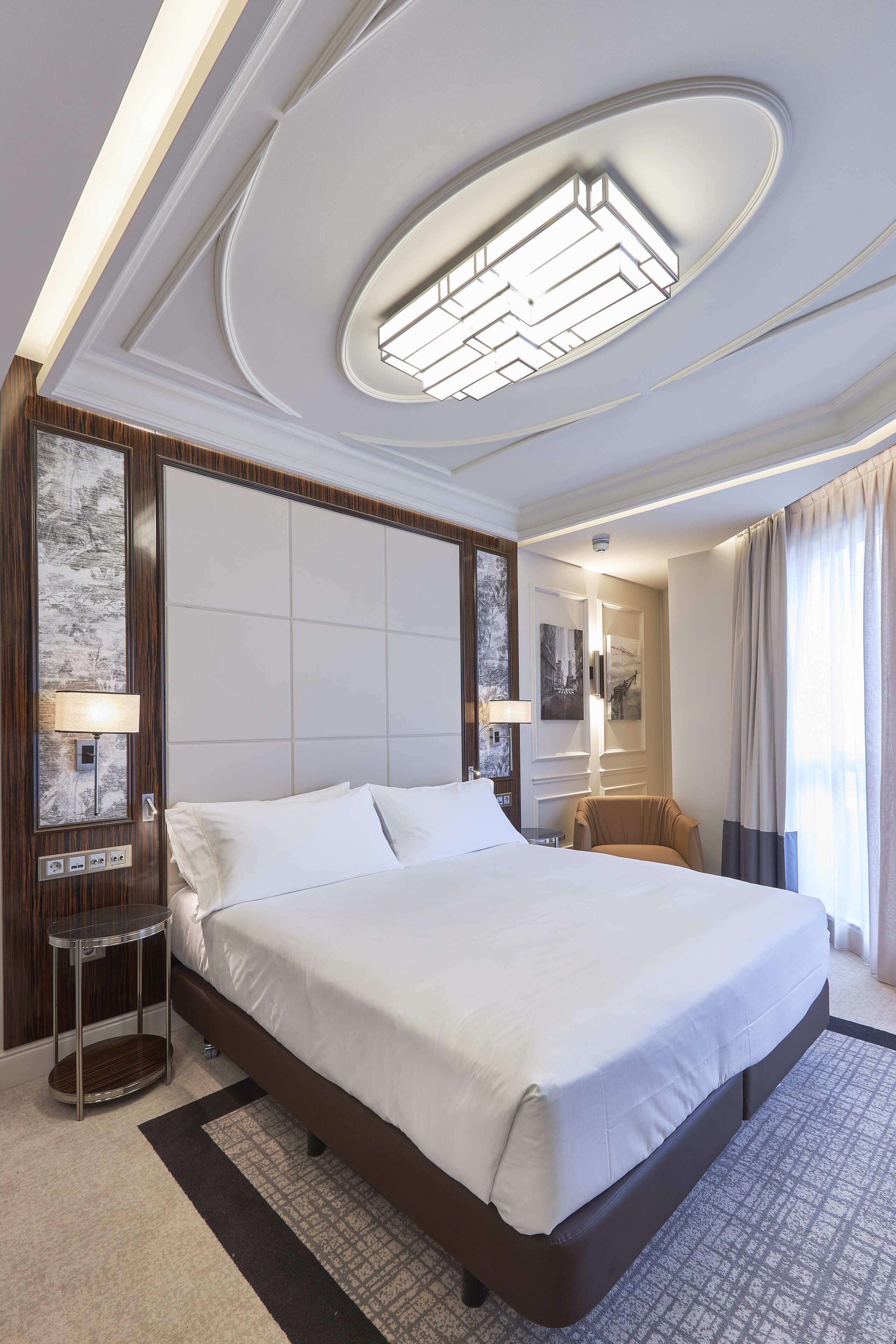 Quarto de hotel moderno com uma cama grande, uma luminária redonda no teto, uma mesinha de cabeceira e um espelho.