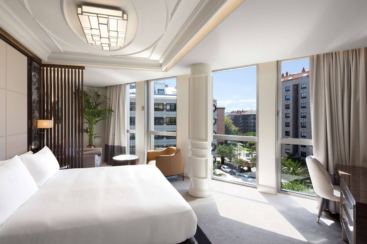 Quarto de hotel com cama grande, escrivaninha, cadeira, planta em vaso e vista da cidade através de grandes janelas.