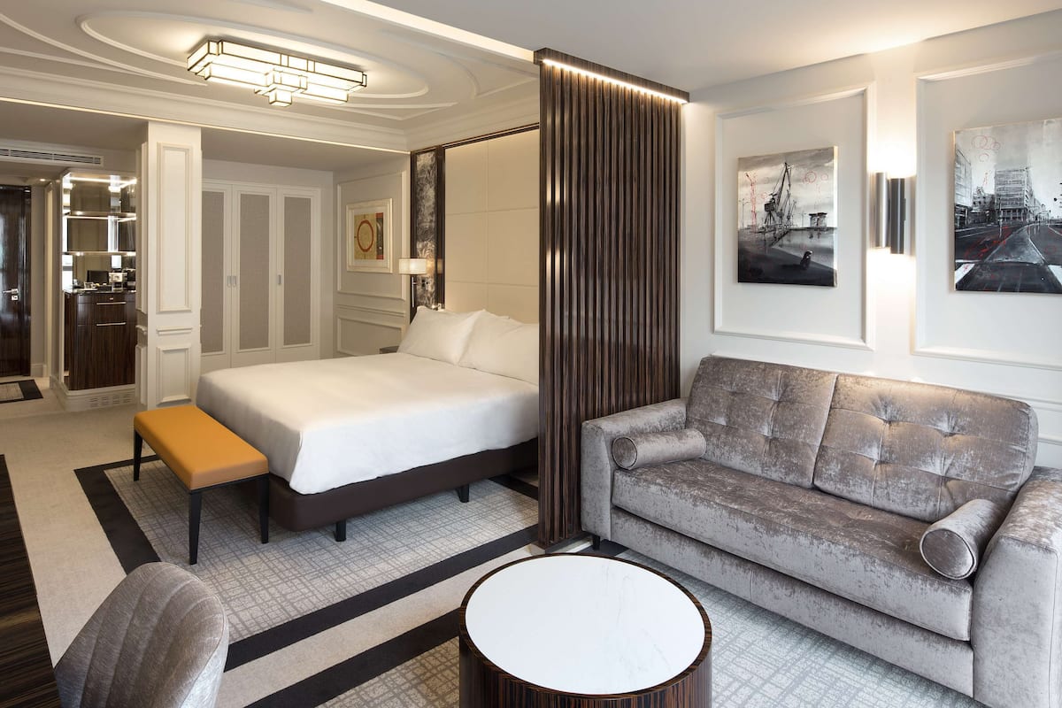 Quarto de hotel moderno com cama de dossel, sofá cinza, mesa de centro redonda e obras de arte nas paredes.
