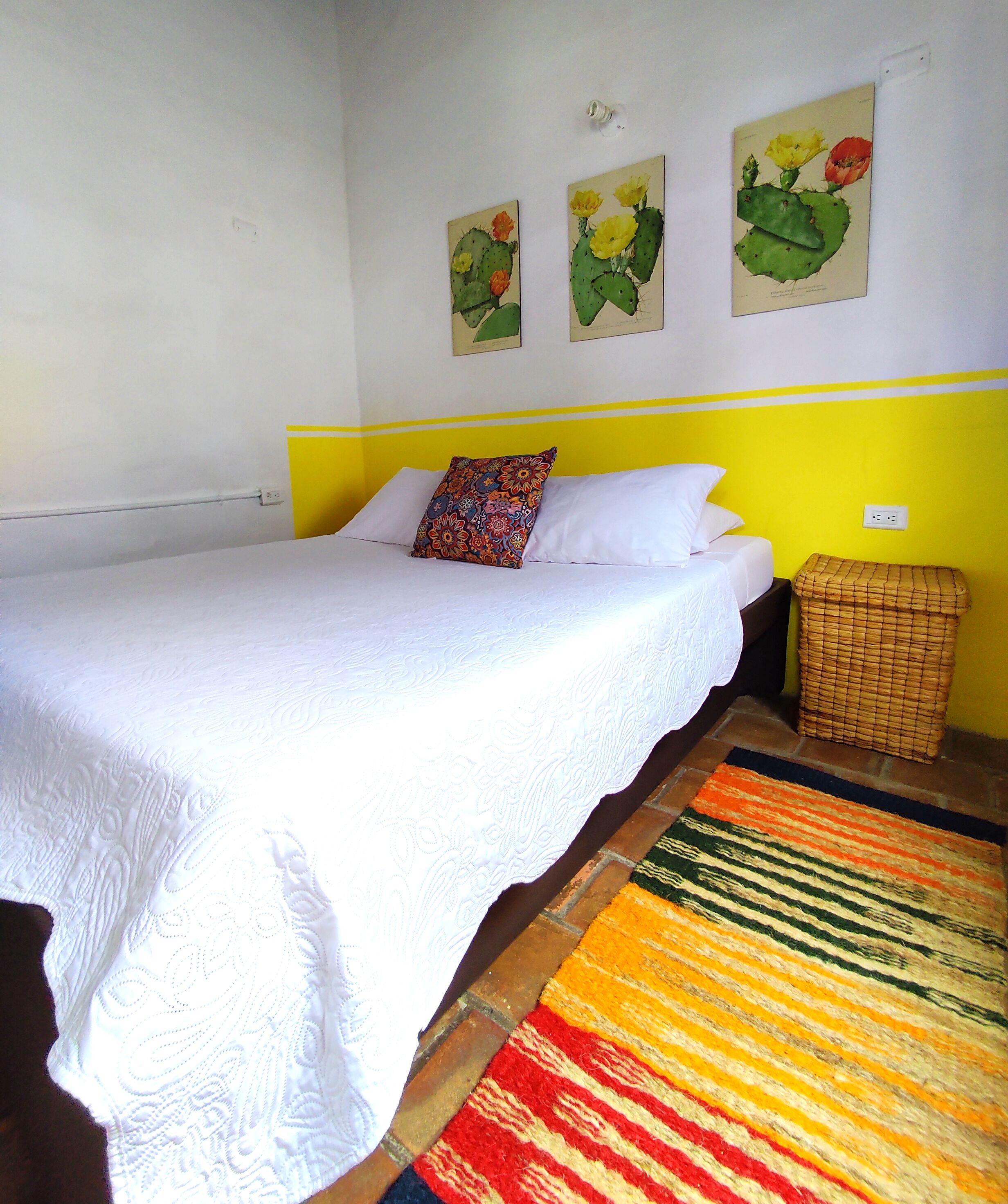 Uma cama com roupa de cama branca, um travesseiro colorido e um tapete estampado. Há três quadros na parede acima da cama.