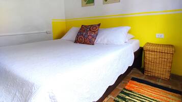 Uma cama com roupa de cama branca, um travesseiro colorido e um tapete estampado. Há três quadros na parede acima da cama.