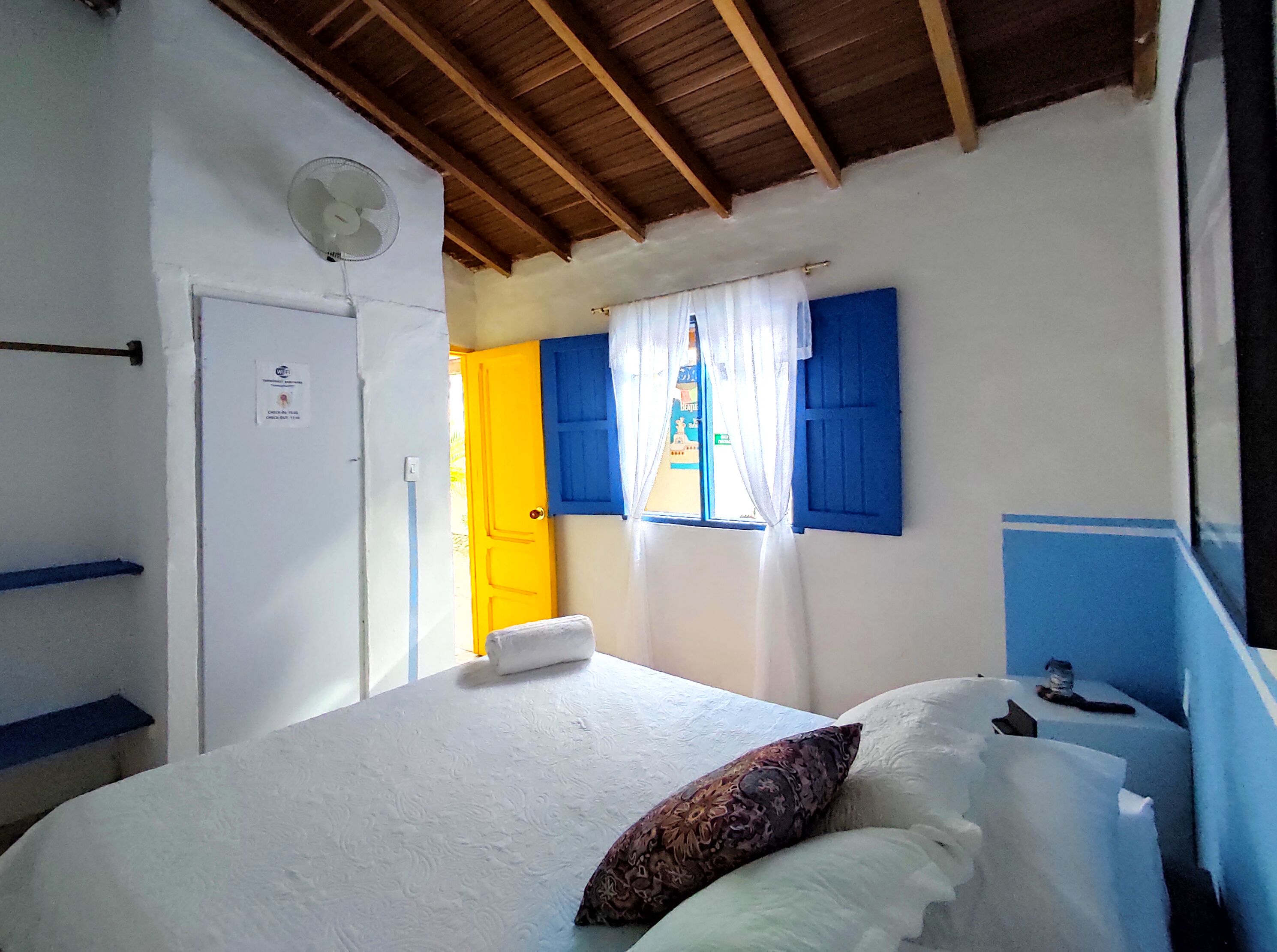 Quarto com cama, mesa de cabeceira, janela com persianas azuis, porta branca e ventilador de teto.