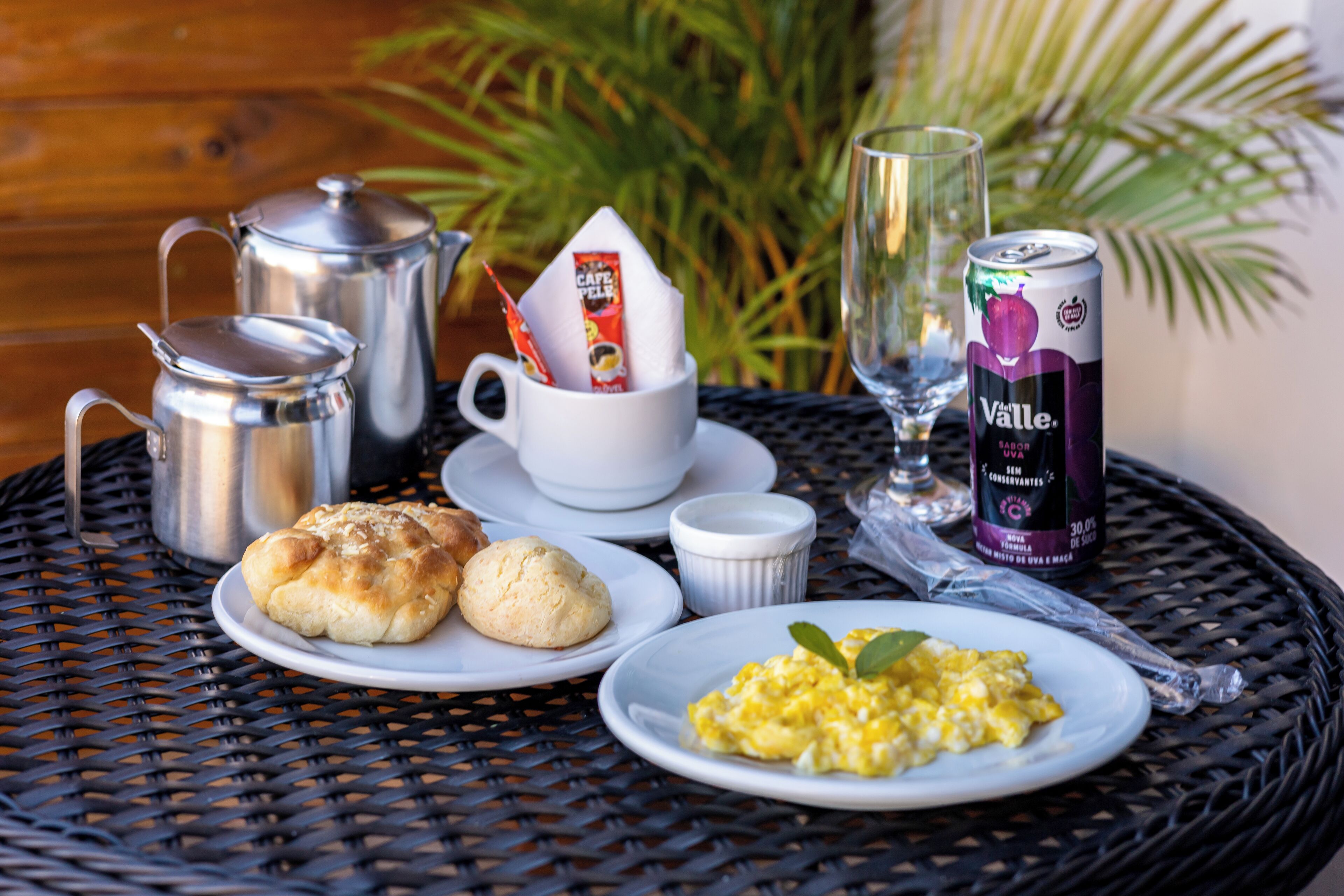 Uma mesa de vime com um conjunto de café da manhã, incluindo cafeteira, bule, croissants, ovos mexidos, uma lata de Valla e um copo de água.