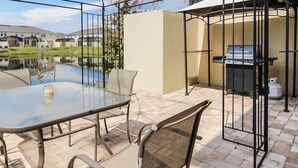 Um pátio externo com mesa e cadeiras de vidro, churrasqueira e vista para um lago e edifícios ao fundo.