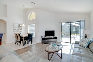 Uma sala de estar ampla com sofá, mesa de centro de vidro, área de jantar com cadeiras, TV e uma grande porta de correr de vidro que dá para uma área externa.