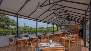 Uma área de refeições ao ar livre coberta, com mesas, cadeiras, janelas amplas e vista para o verde.