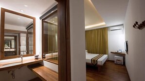 Quarto de hotel com toucador de madeira, um espelho grande, cama com cabeceira, televisão e pequena mesa com abajur.