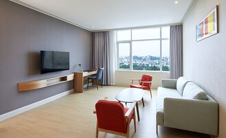 Quarto de hotel moderno com televisão de tela plana na parede, escrivaninha com cadeira, sofá e uma grande janela com vista para a cidade.