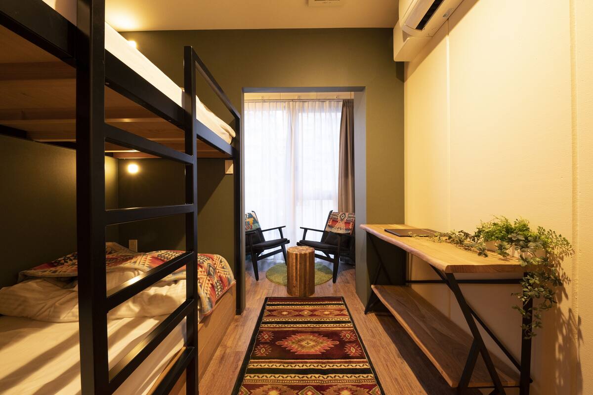 Quarto compacto com beliches, uma pequena mesa, duas cadeiras e uma janela com cortinas.