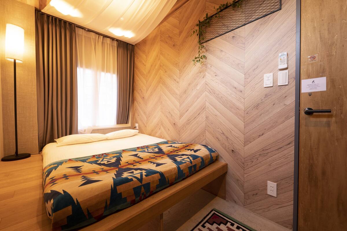 Quarto de hotel com cama, mesa de cabeceira, luminária, janela com cortinas e parede de madeira com padrão herringbone.