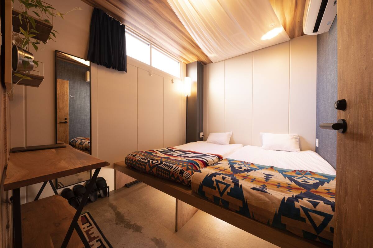 Quarto compacto com cama, uma mesinha, um espelho e janela com cortinas.