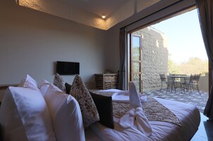 Quarto com cama grande, almofadas, televisão de tela plana e vista para um pátio externo com mesa e cadeiras.