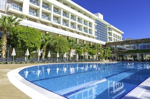 Uma piscina grande com espreguiçadeiras e guarda-sóis, cercada por palmeiras e um edifício de hotel com vários andares e varandas.