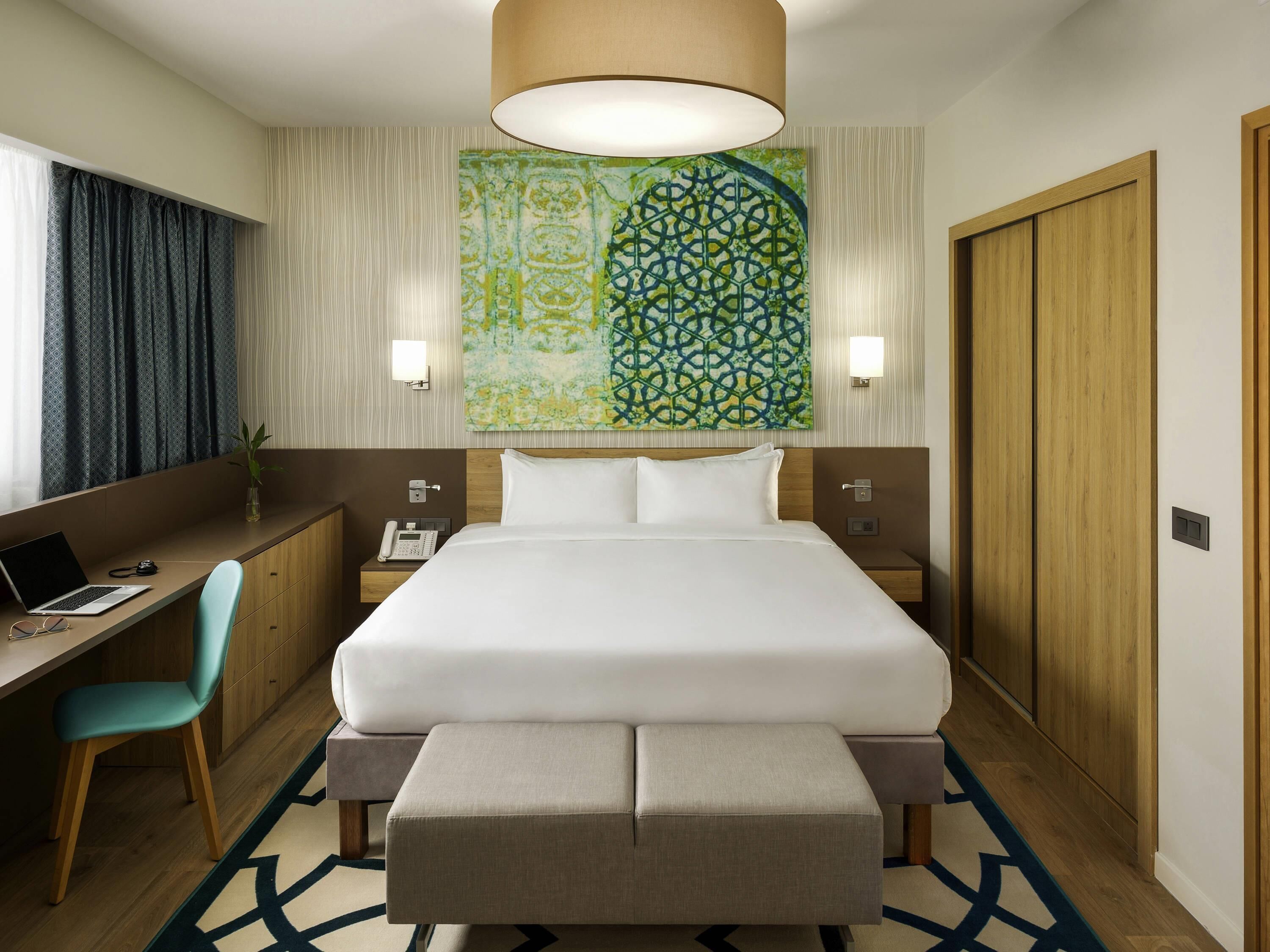 Quarto de hotel com cama, escrivaninha, cadeira, mesas de cabeceira, abajur, obras de arte e guarda-roupa.