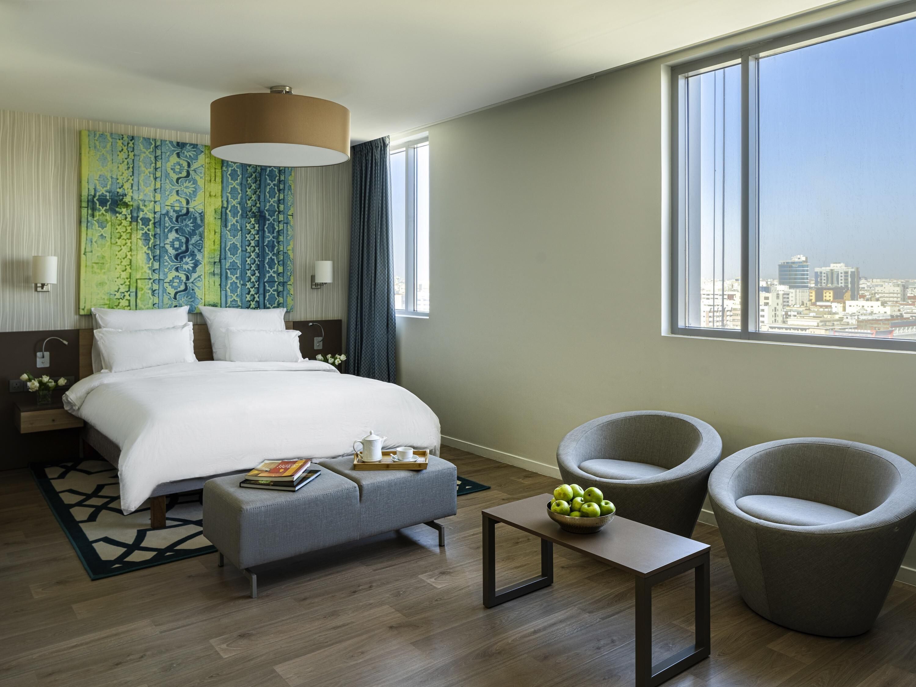 Quarto de hotel moderno com uma cama grande, duas poltronas, uma mesa pequena e vista para a cidade.