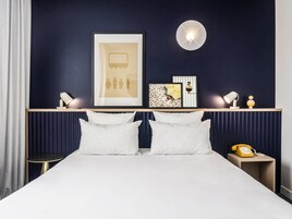 Quarto de hotel com cama, duas mesas de cabeceira, criado-mudo com telefone, luminária na parede e quadro acima da cama.