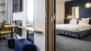 Quarto de hotel com uma cama, uma secretária com cadeira, uma televisão e um espelho.