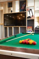 Uma mesa de bilhar com bolas, uma televisão e prateleiras com livros e um skate.