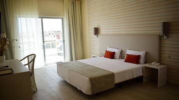 Quarto de hotel com uma cama grande, uma escrivaninha e uma cadeira. Há uma janela com cortinas, uma parede estampada e uma mesa de cabeceira com um telefone.