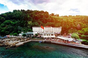 Um hotel à beira-mar com um edifício grande, um cais com barcos e uma encosta arborizada ao fundo.