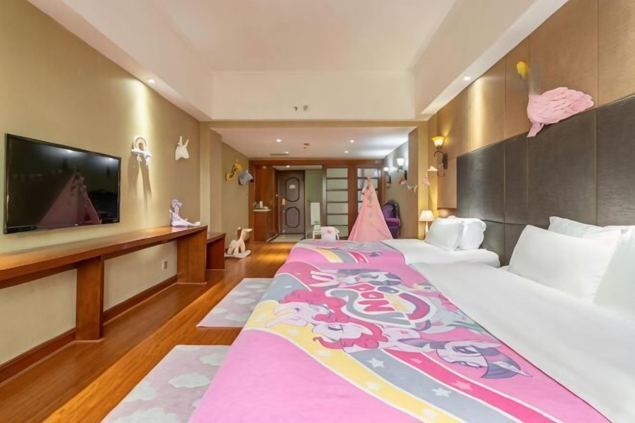 Quarto de hotel com cama, televisão, escrivaninha de madeira e edredom rosa e cinza.