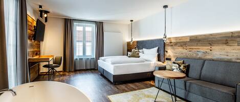 Quarto de hotel moderno com uma banheira grande, uma cama com cabeceira de madeira, um sofá cinzento e uma pequena mesa de madeira.